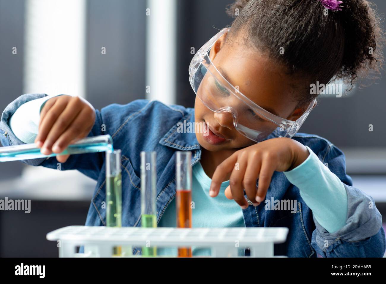 Happy biracial schoolgirl in safety glasses doing experiment in science ...