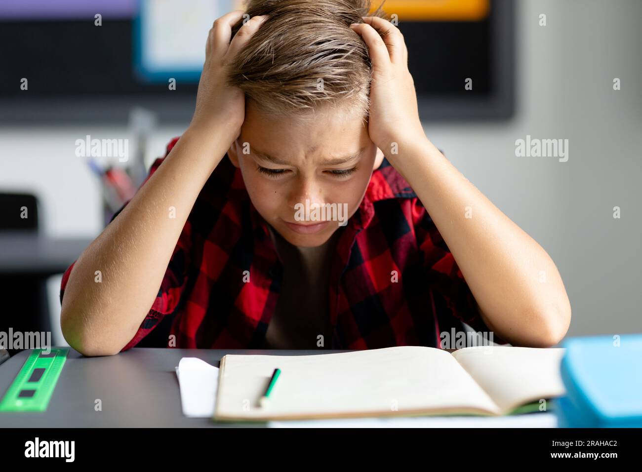 Stressed caucasian schoolboy at desk, head in hands, staring at book in ...