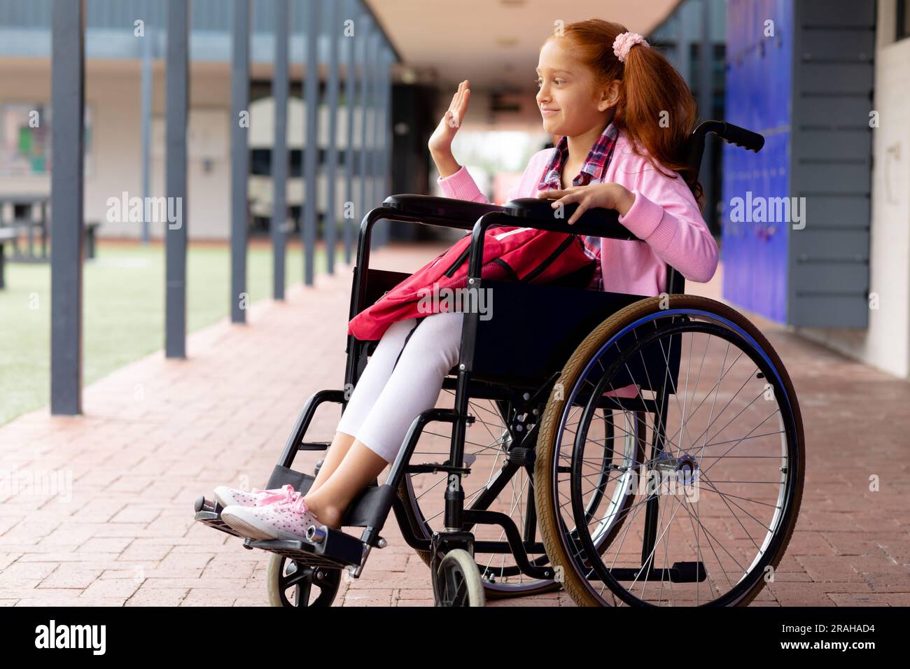 Happy biracial schoolgirl in wheelchair, waving in school corridor ...