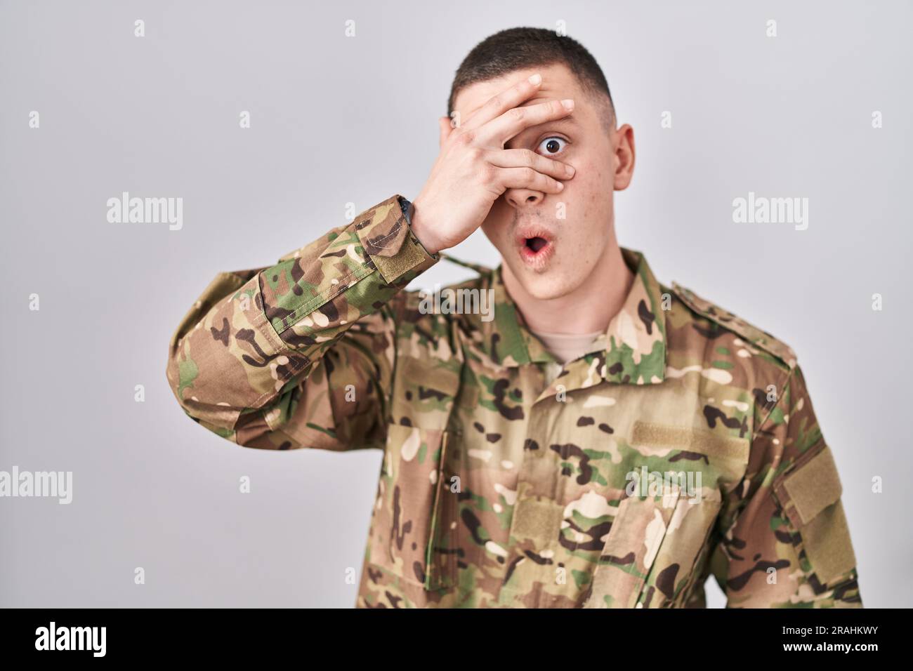 Young man wearing camouflage army uniform peeking in shock covering ...