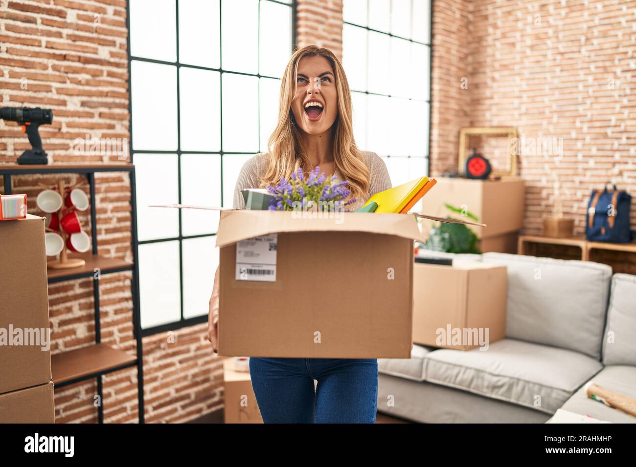 Young blonde woman moving to a new home holding cardboard box angry and ...