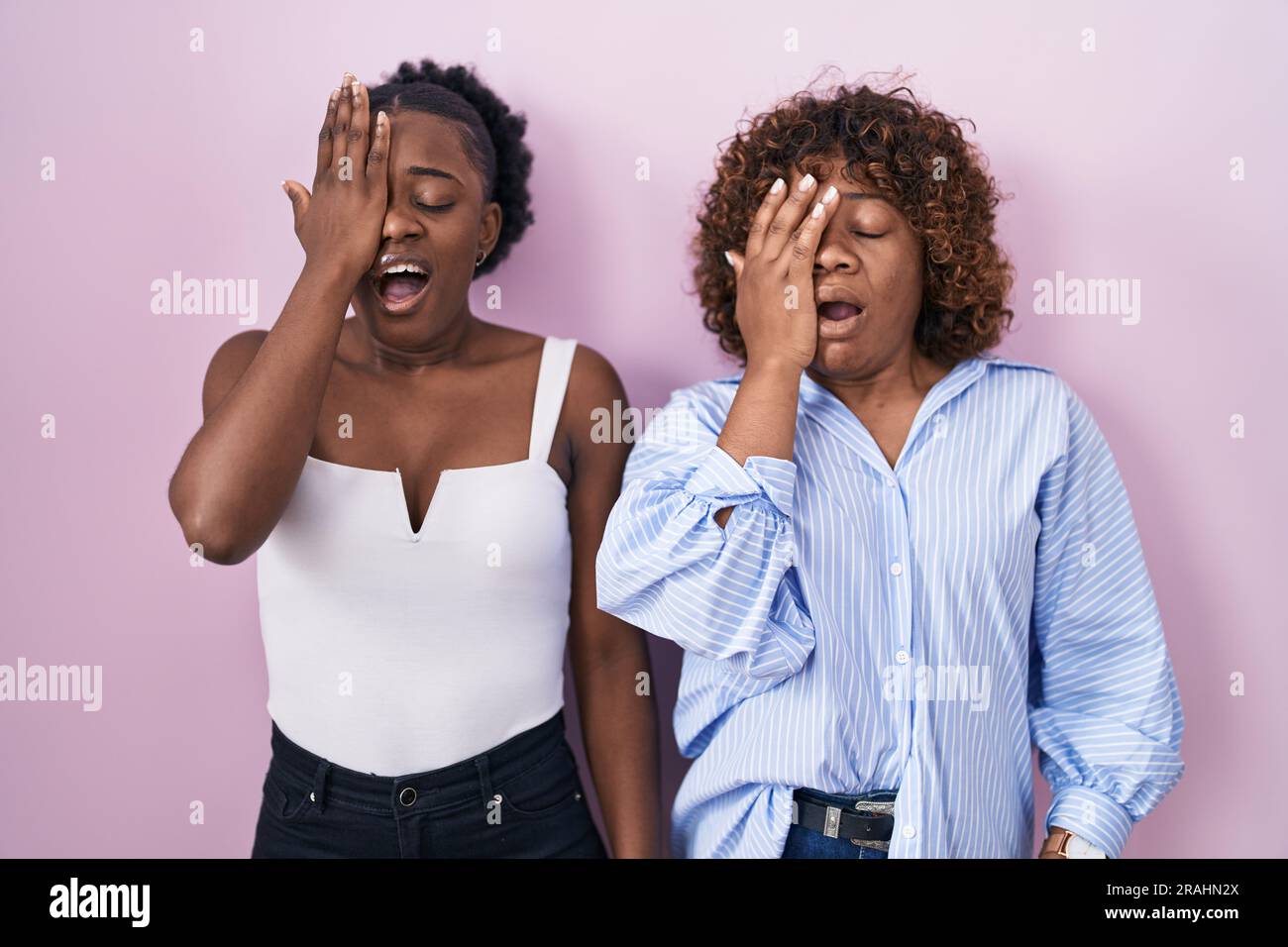 Two african women standing over pink background yawning tired covering ...