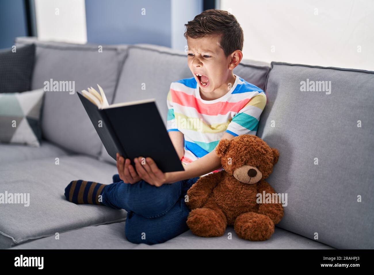 Young caucasian kid sitting on the sofa reading a book angry and mad ...