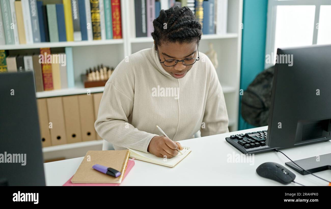 African american woman student using computer writing notes at library ...