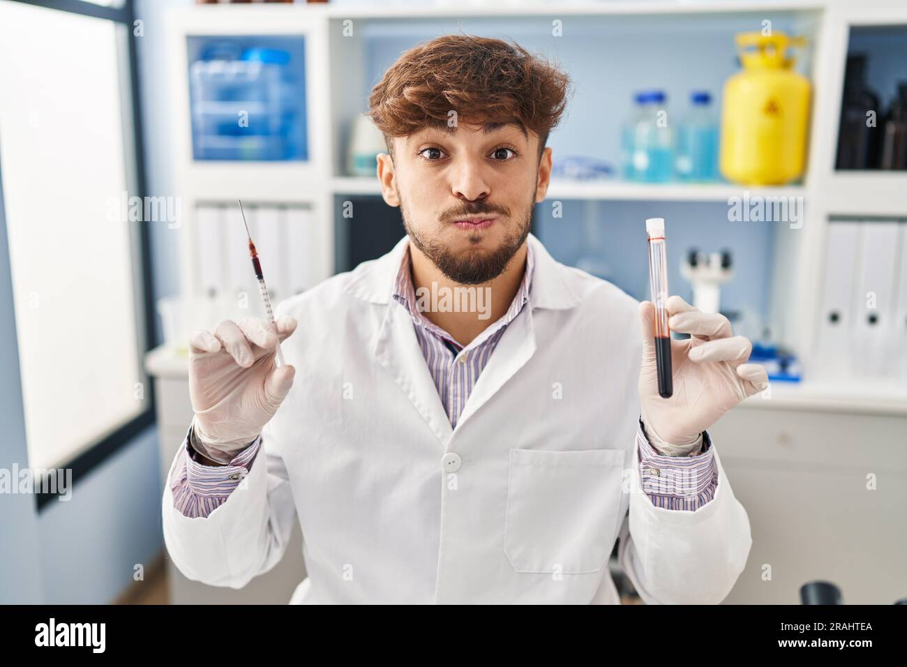 Arab man with beard working at scientist laboratory holding blood sample puffing cheeks with ...