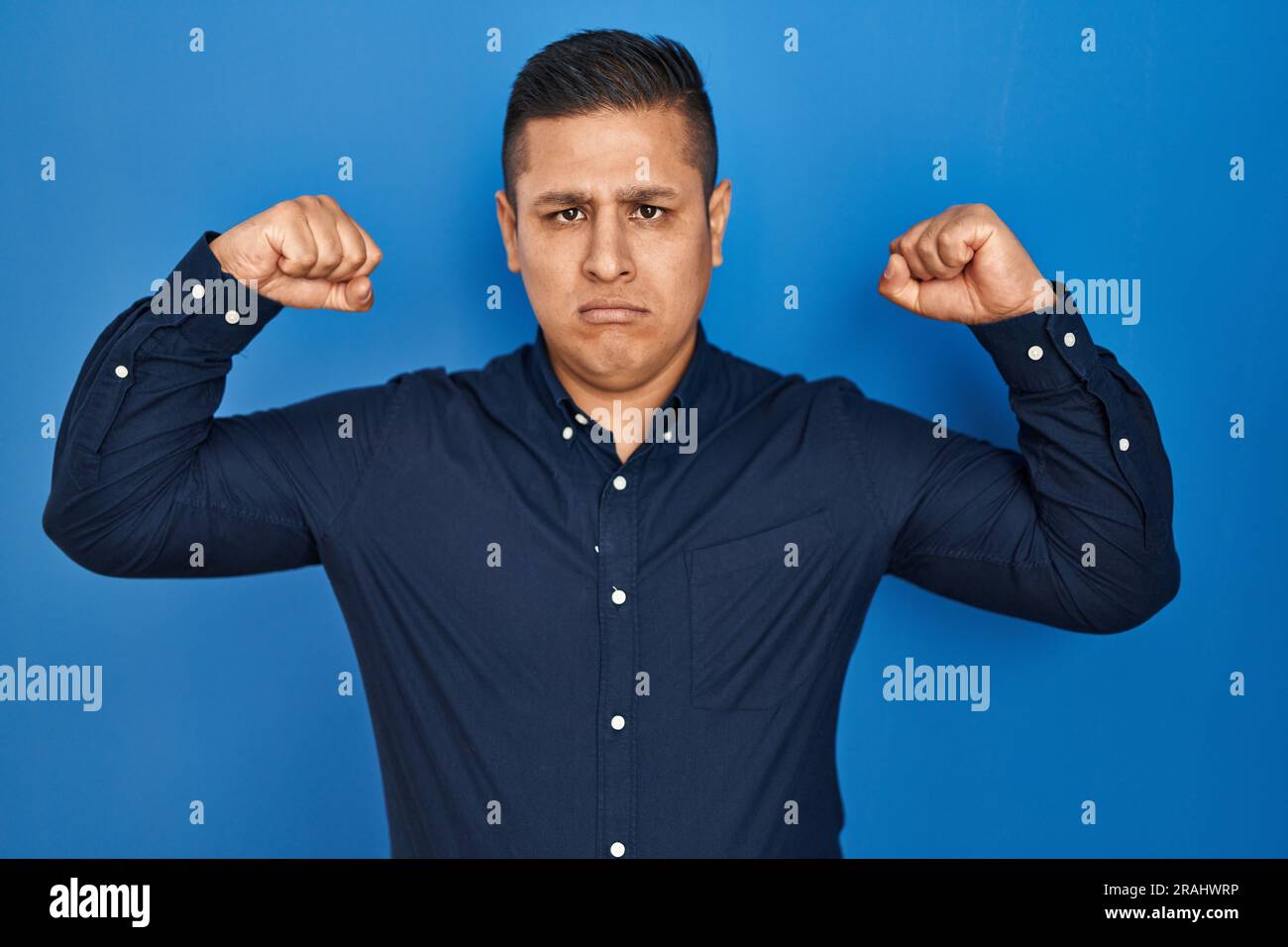 Hispanic Young Man Standing Over Blue Background Showing Arms Muscles Smiling Proud Fitness