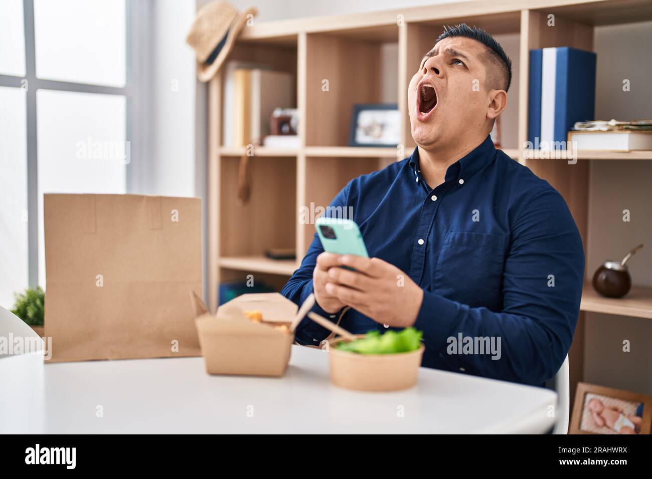 Hispanic young man eating take away food using smartphone angry and mad ...