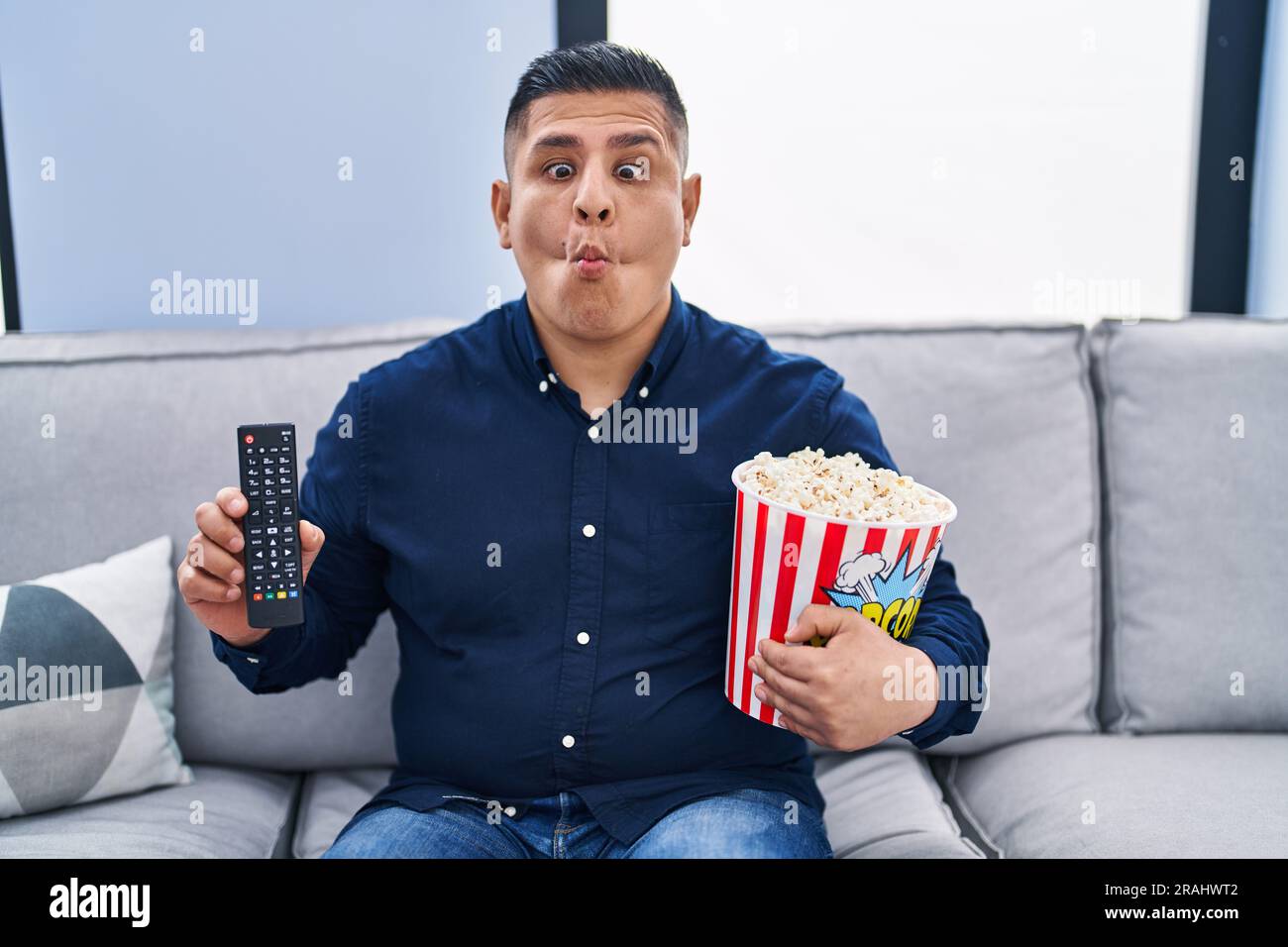 Hispanic young man eating popcorn using tv control making fish face ...