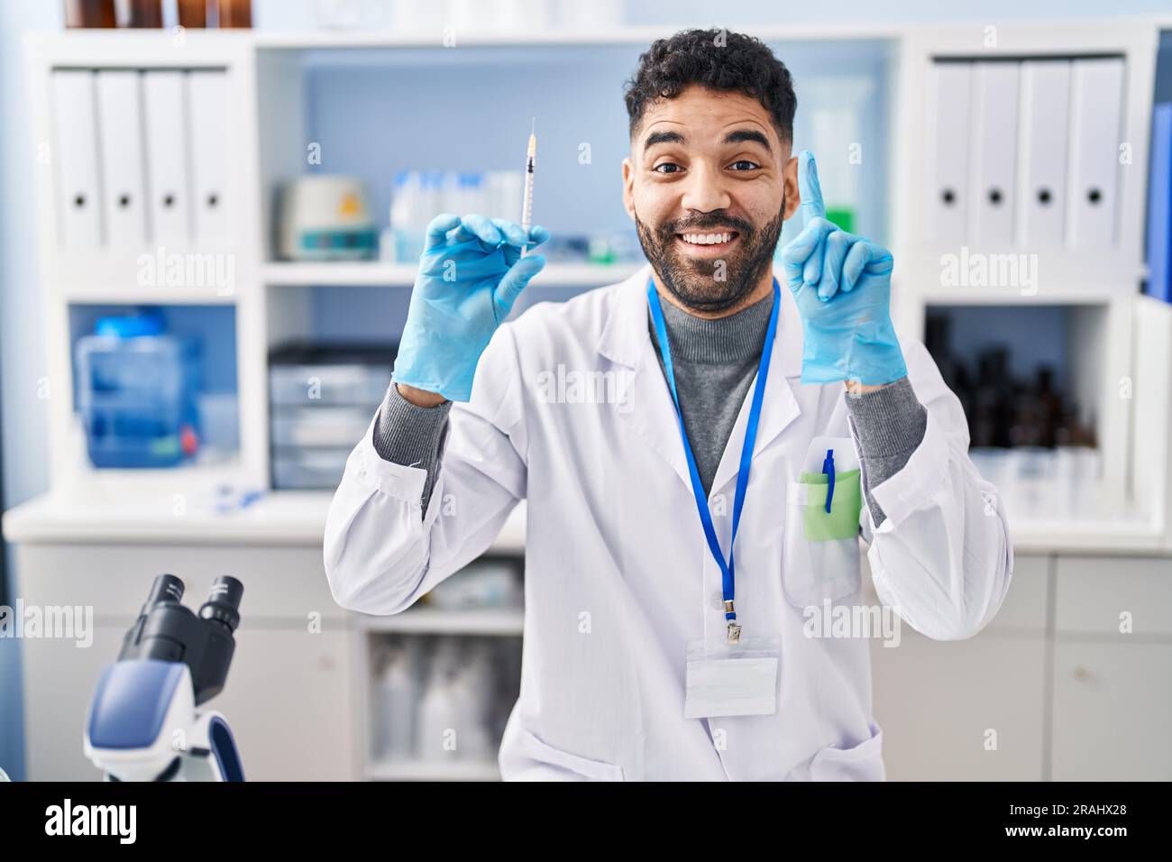 Hispanic man with beard working at scientist laboratory holding syringe smiling with an idea or ...