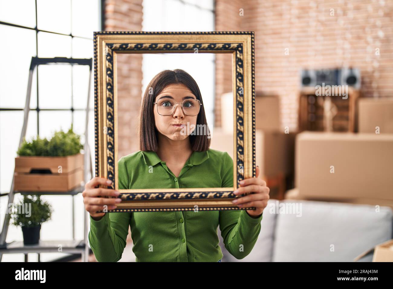 Young hispanic woman at new home holding empty frame puffing cheeks ...