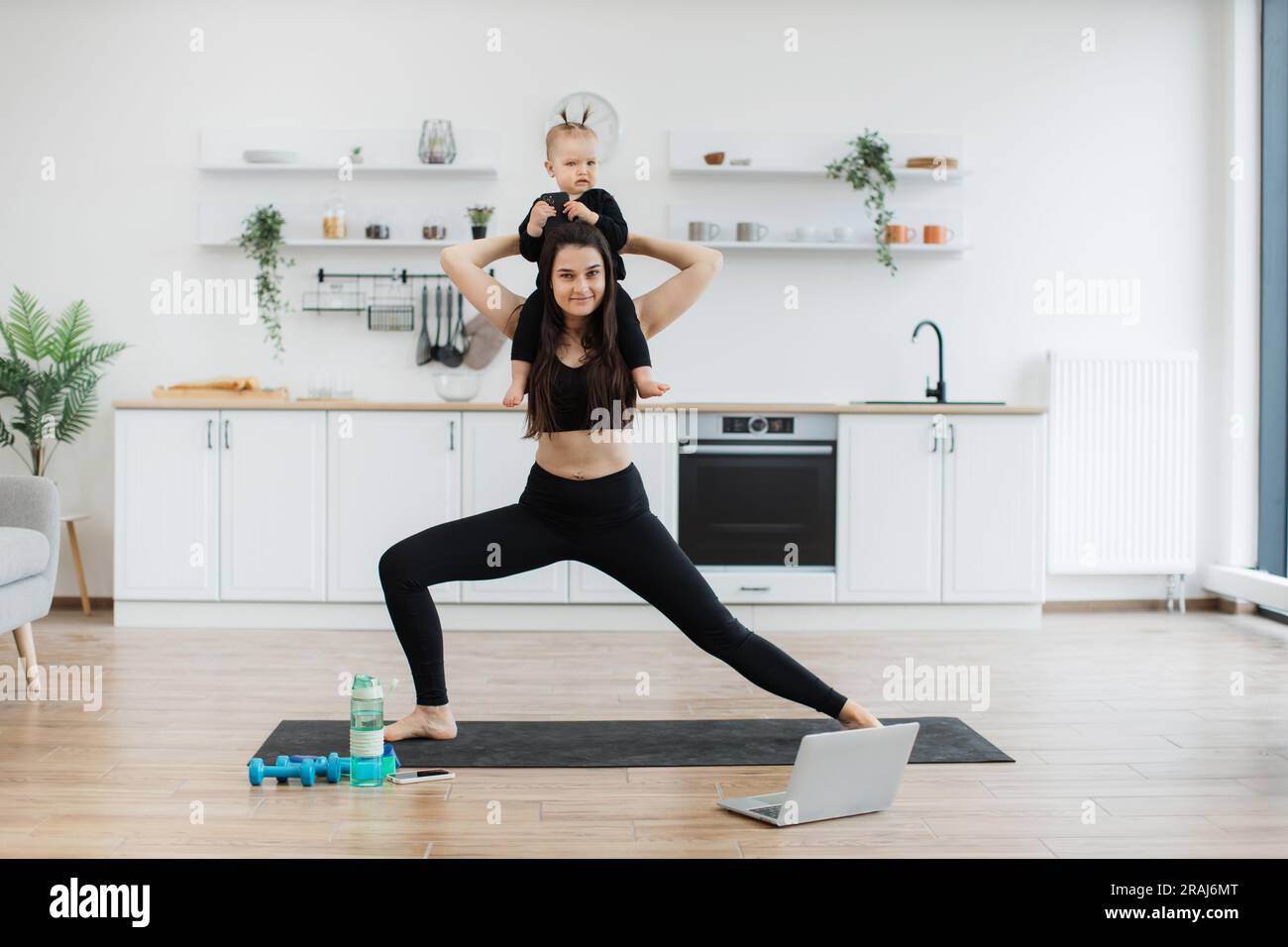 Sporty female keeping little girl on shoulders while stretching knees in standing position on ...