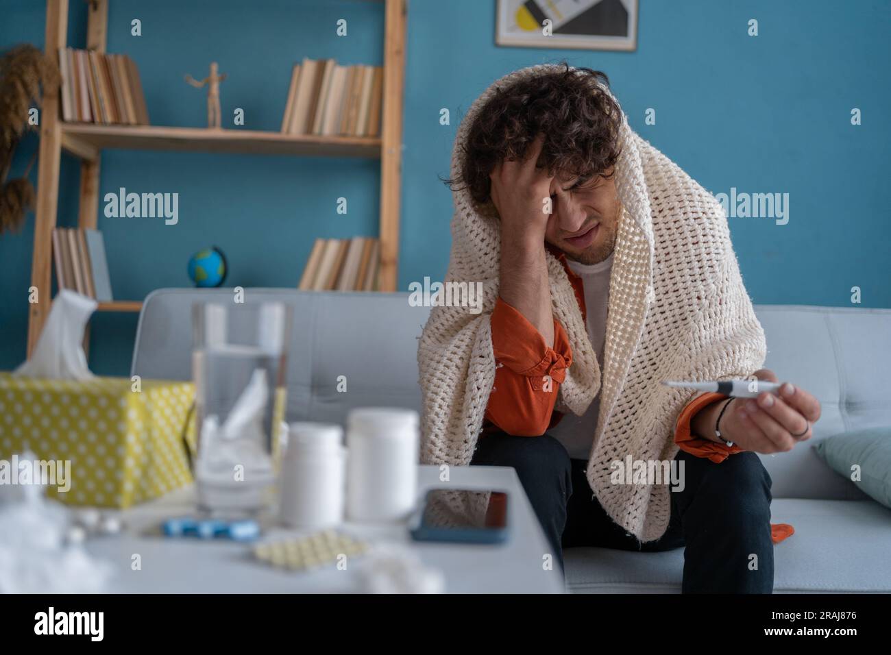 Sick man, wrapped in a blanket measuring a temperature while sitting on ...