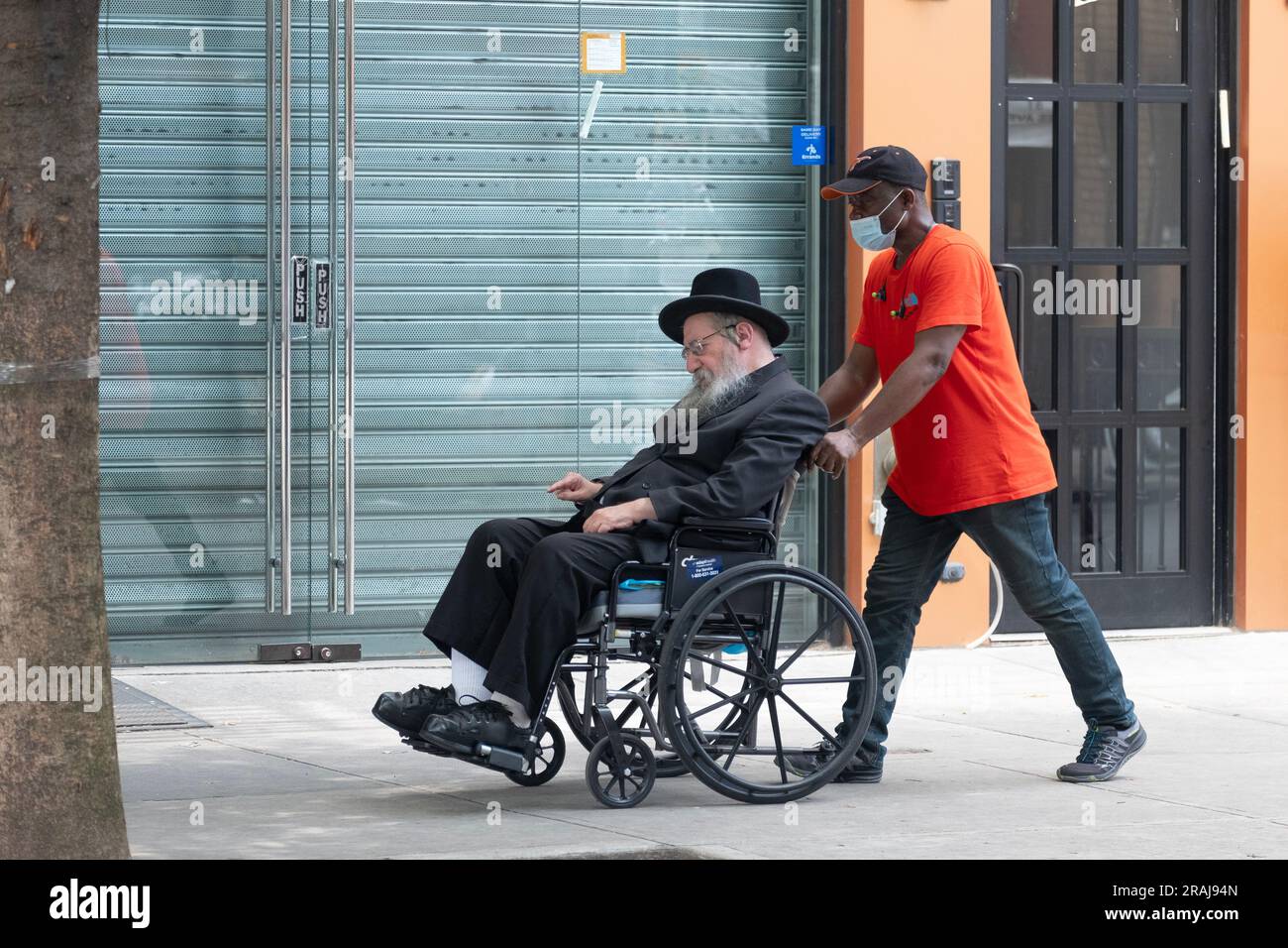 A senior Hasidic Jewish man in his wheelchair assisted by his aide.. On a quiet street in ...