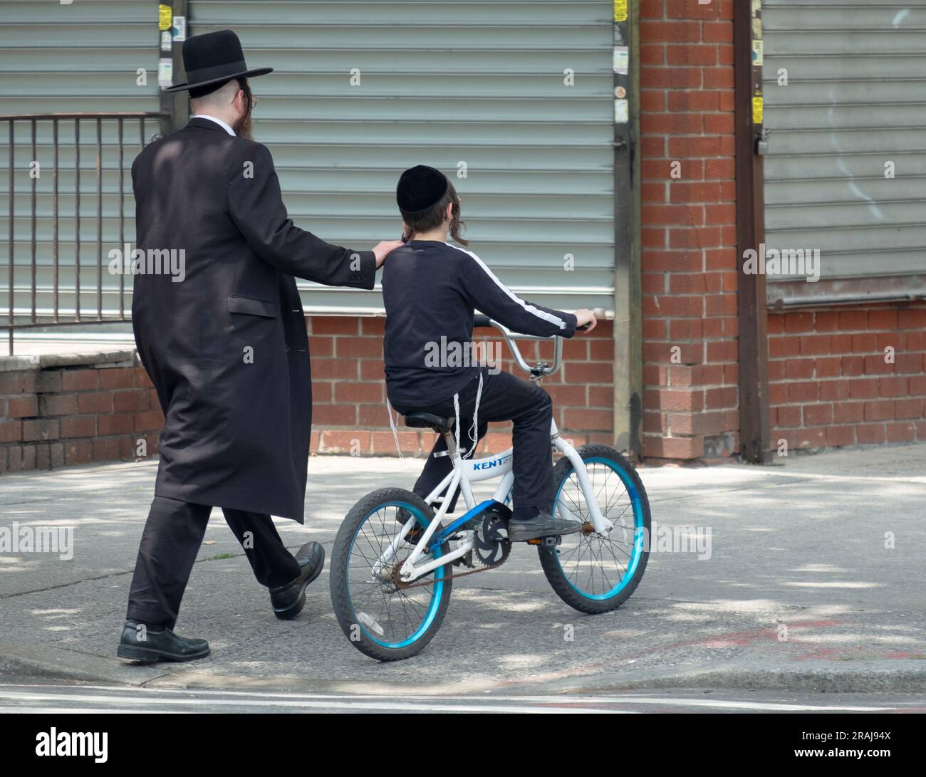 A Hasidic father lends his son support as they walk/bike on a busy ...