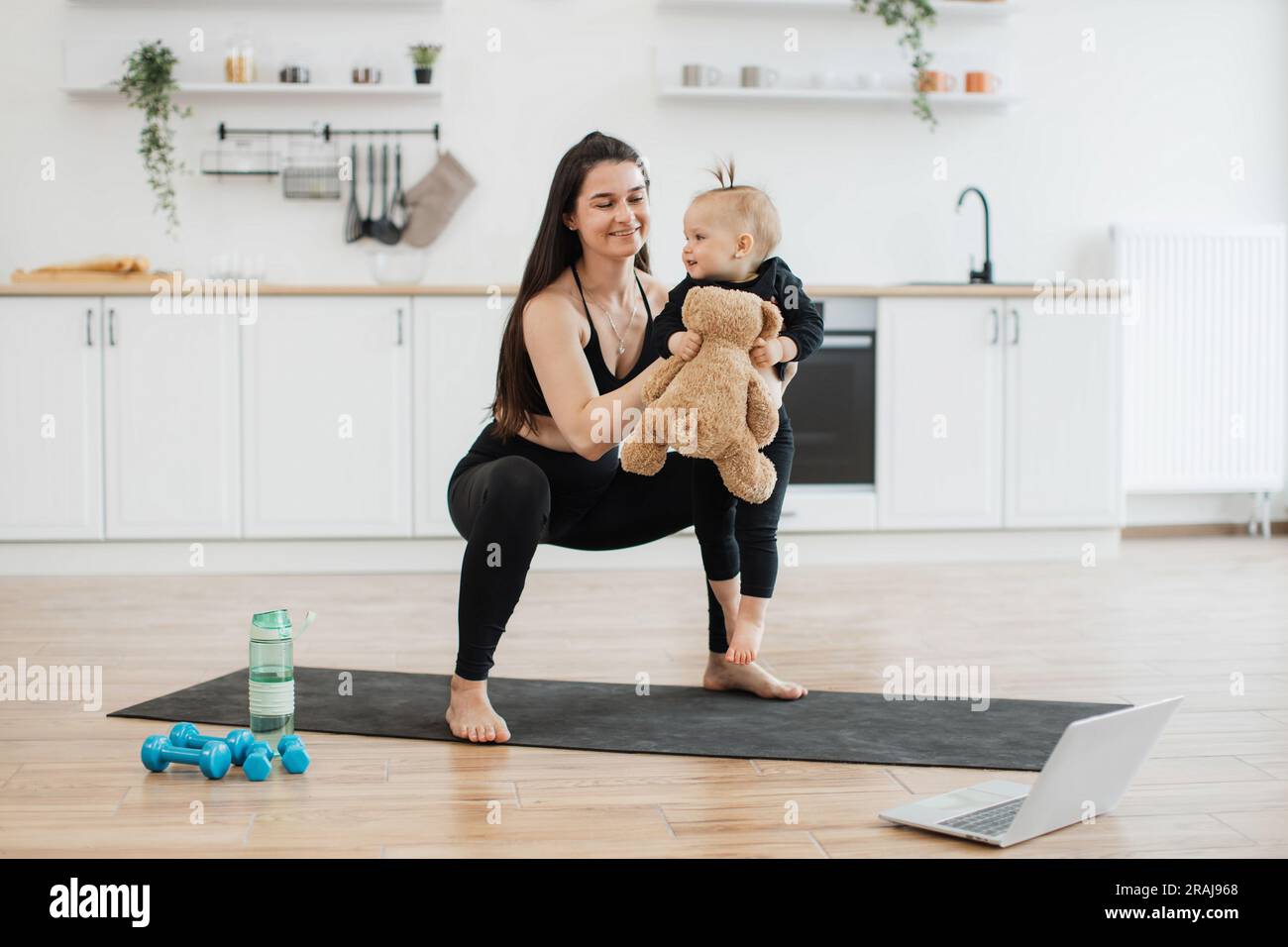 Cheerful person in yoga clothes squatting with infant daughter holding ...
