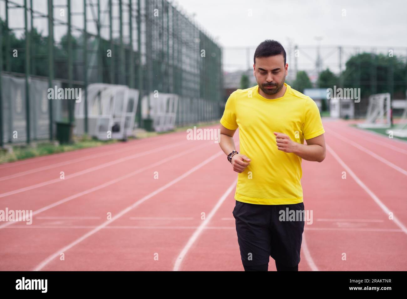 Focused athlete in t-shirt running along red track past soccer field ...