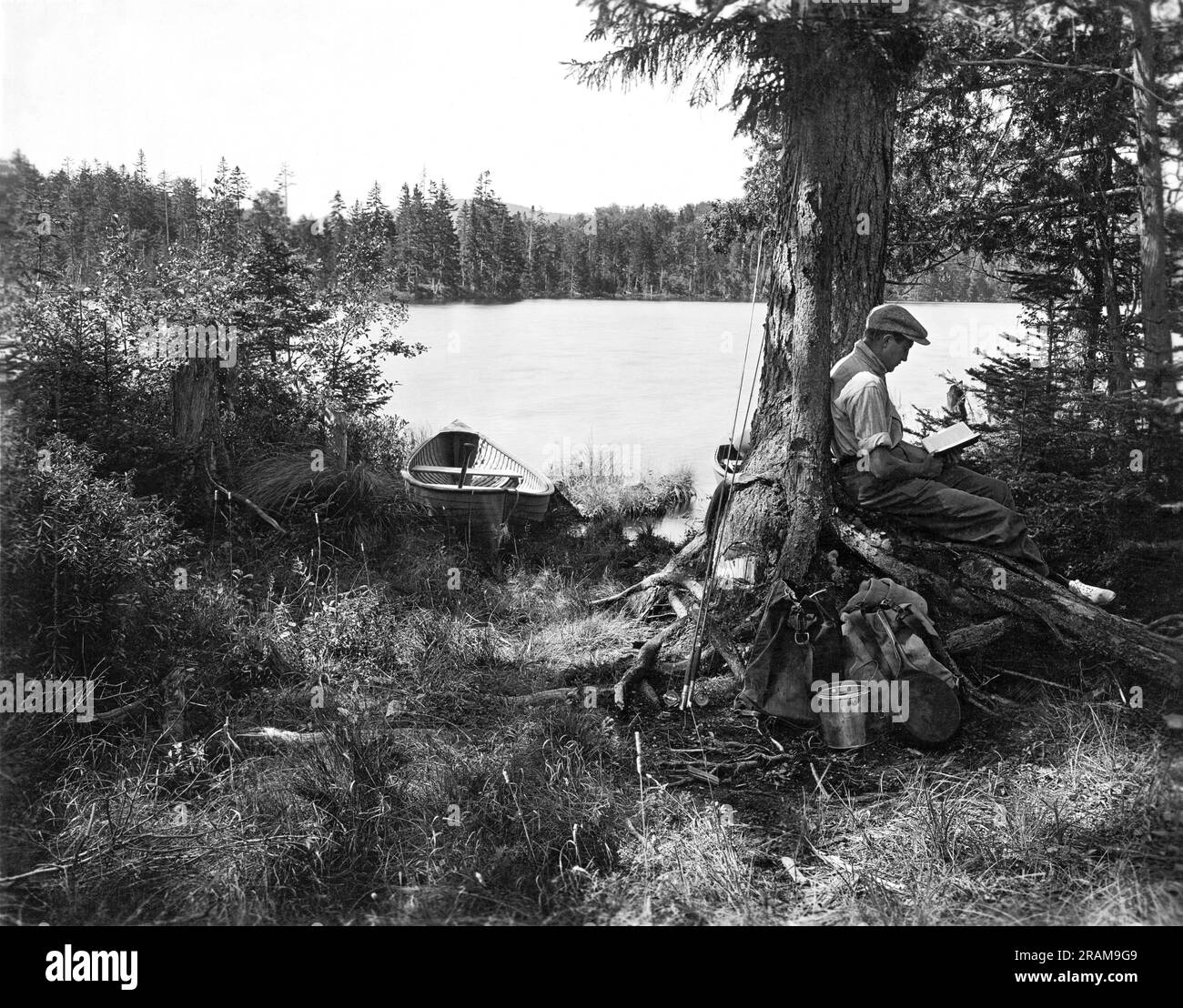 Maine: c. 1923 A solitary man reading a book at his campsite on a lake ...