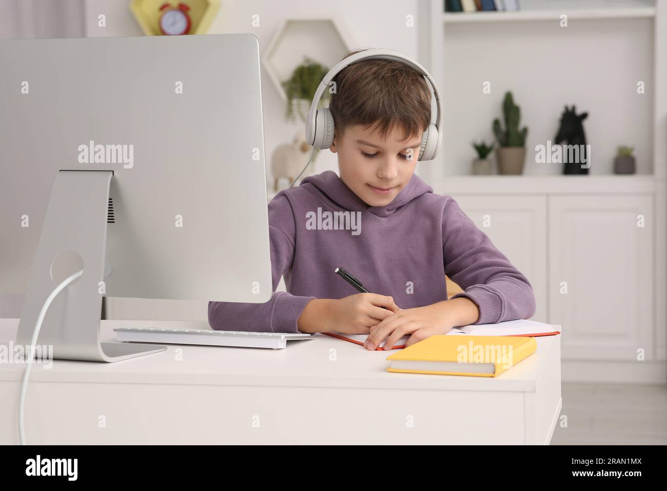 Boy writing in notepad while using computer and headphones at desk in ...
