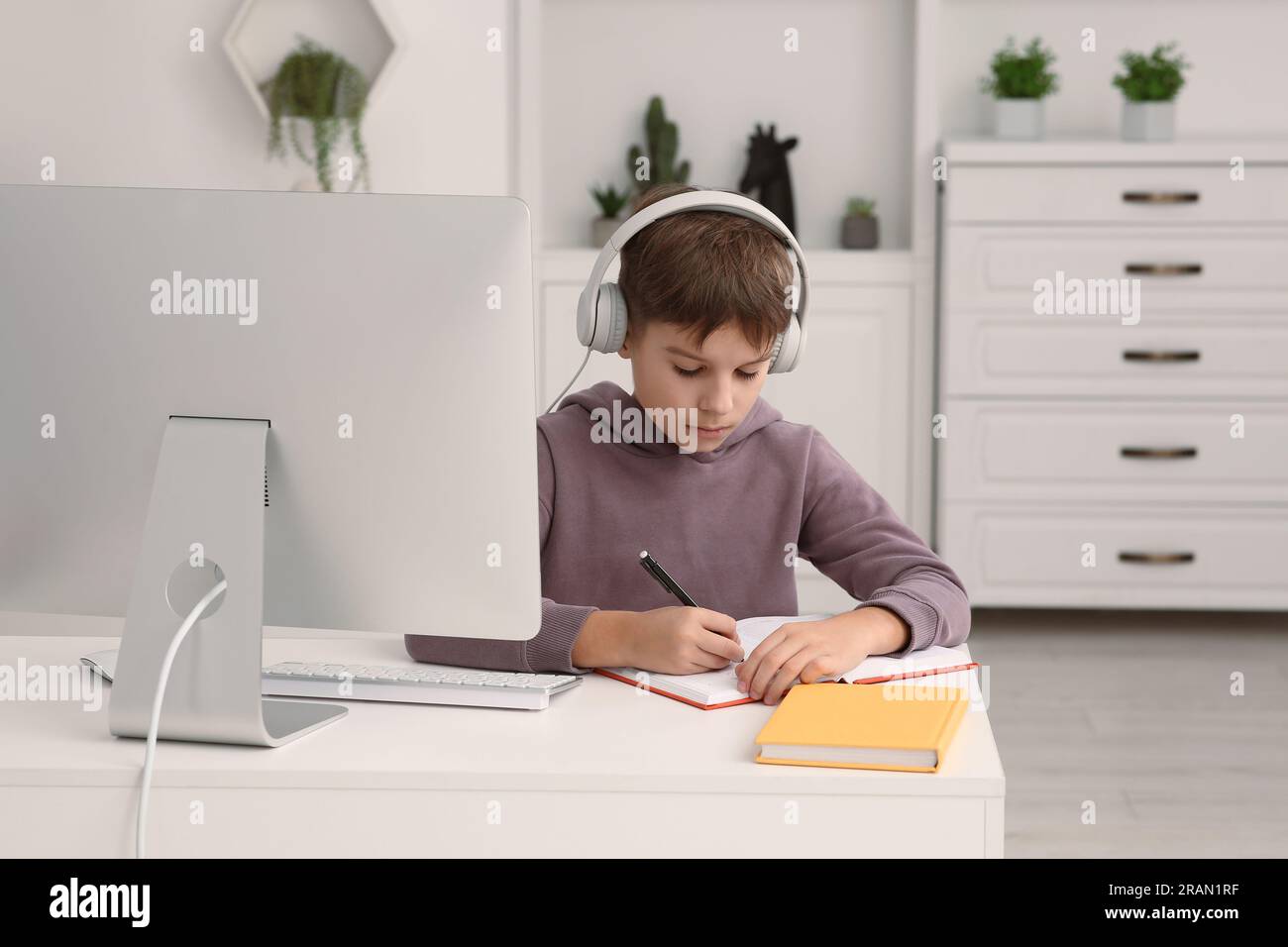 Boy writing in notepad while using computer and headphones at desk in ...