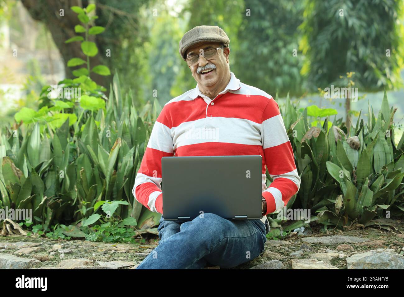 Old man sitting in a lush green atmosphere in a park holding a laptop ...