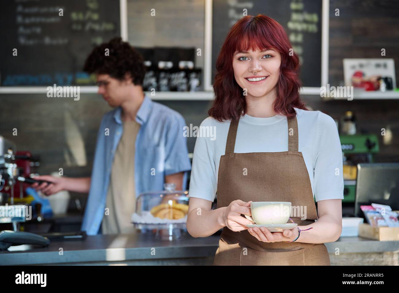 Portrait of young female waitress in front of counter in coffee shop ...