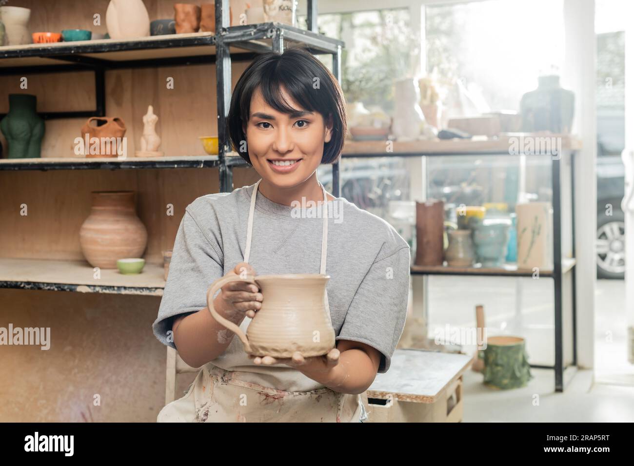 Cheerful young asian female artisan in apron holding clay jug and ...