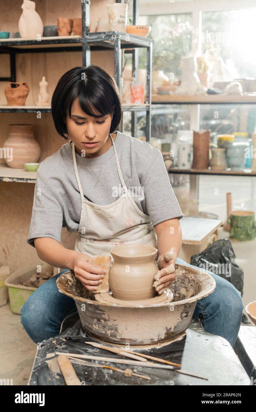 Young brunette asian craftswoman in apron holding scraper near clay ...