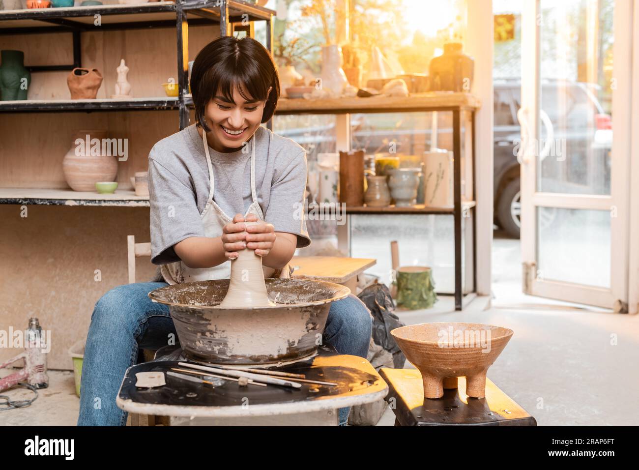 Cheerful young asian female potter in apron molding clay on pottery ...