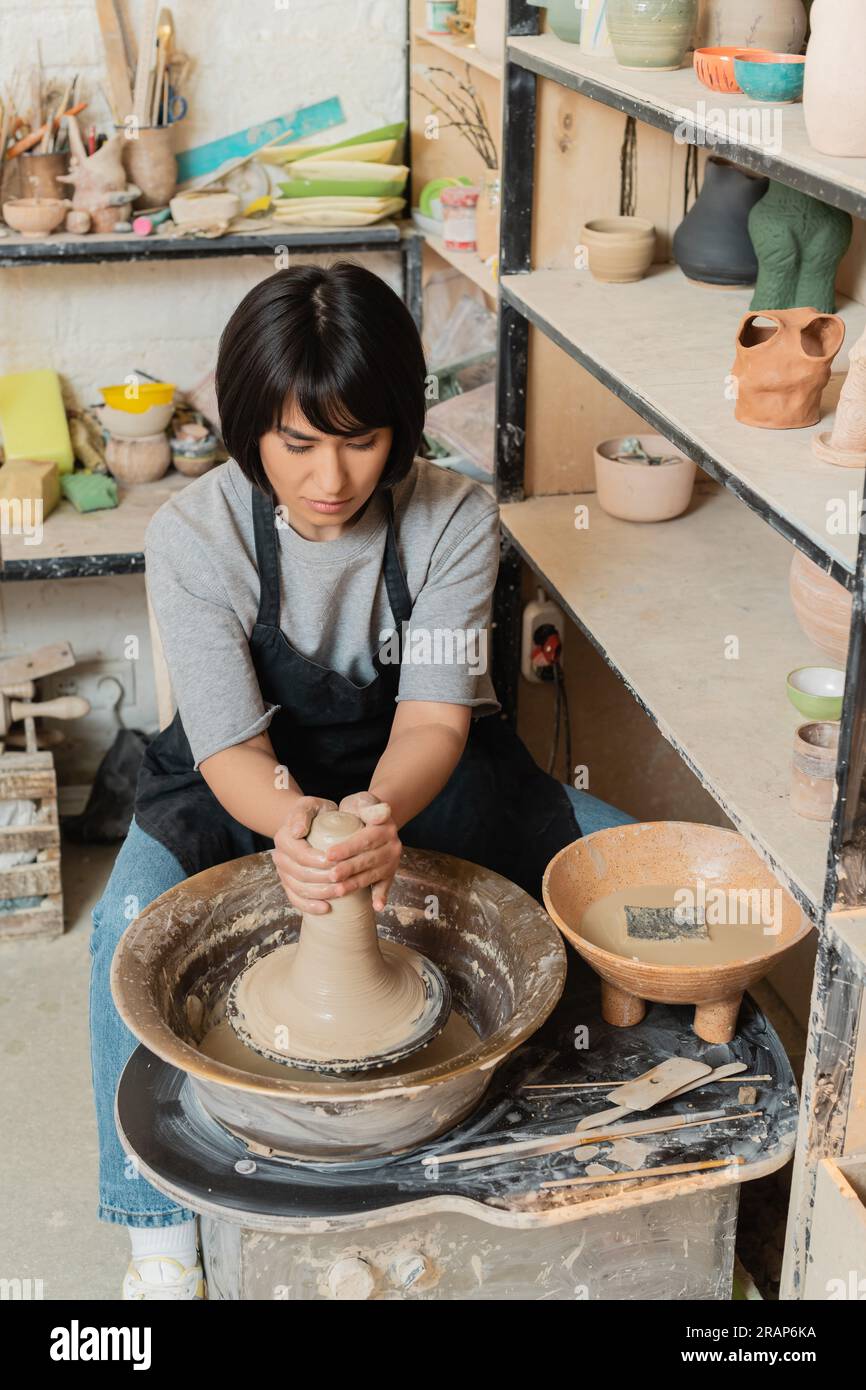 Young asian female artisan in apron molding wet clay on pottery wheel ...