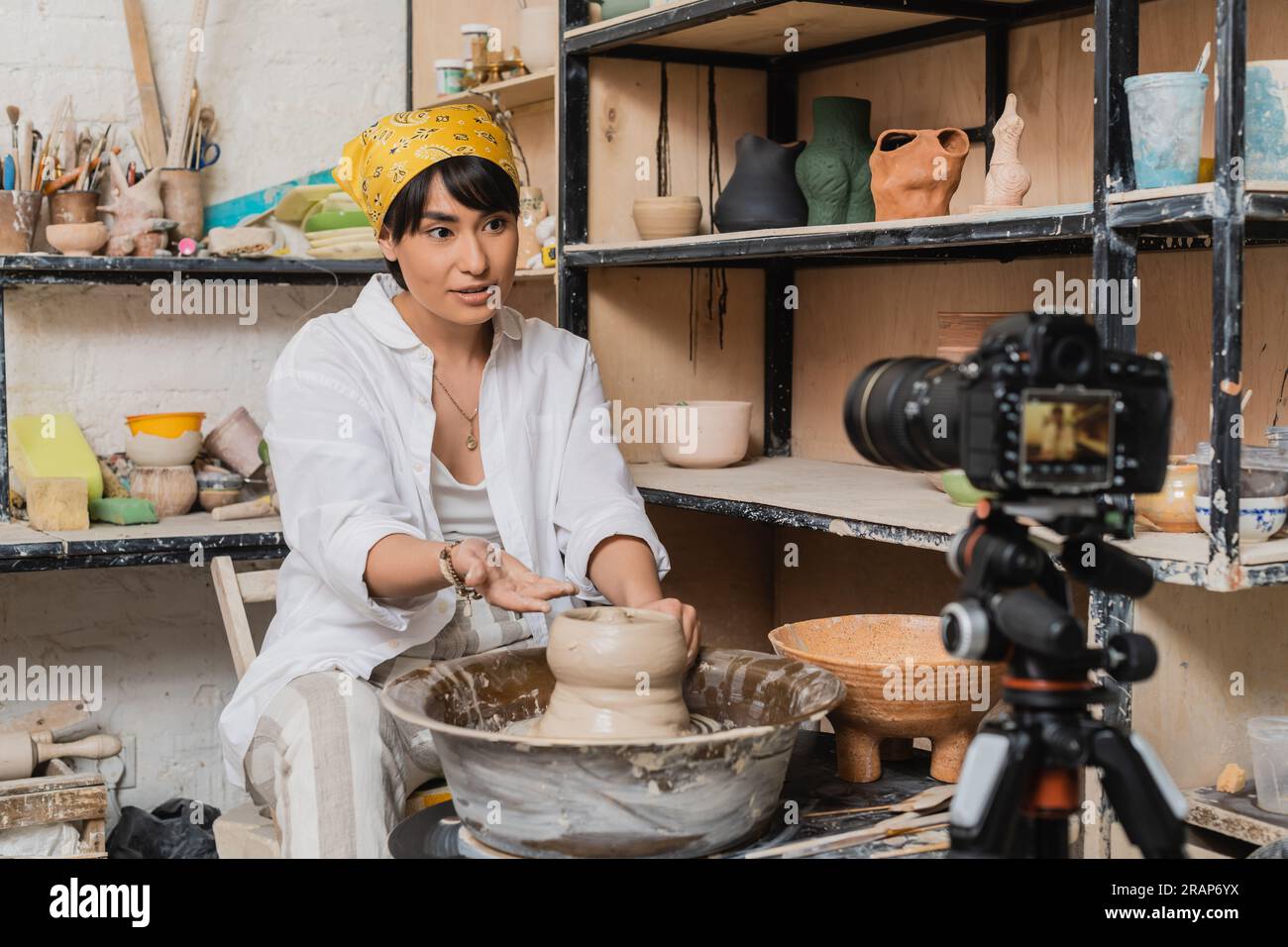 Asian female artisan in workwear and headscarf talking and gesturing at digital camera on tripod ...
