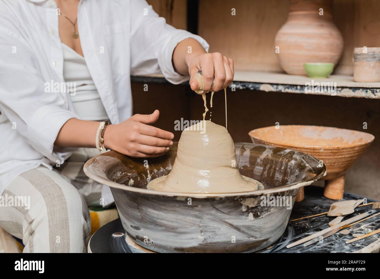 Cropped view of blurred female artist in workwear pouring water on clay ...
