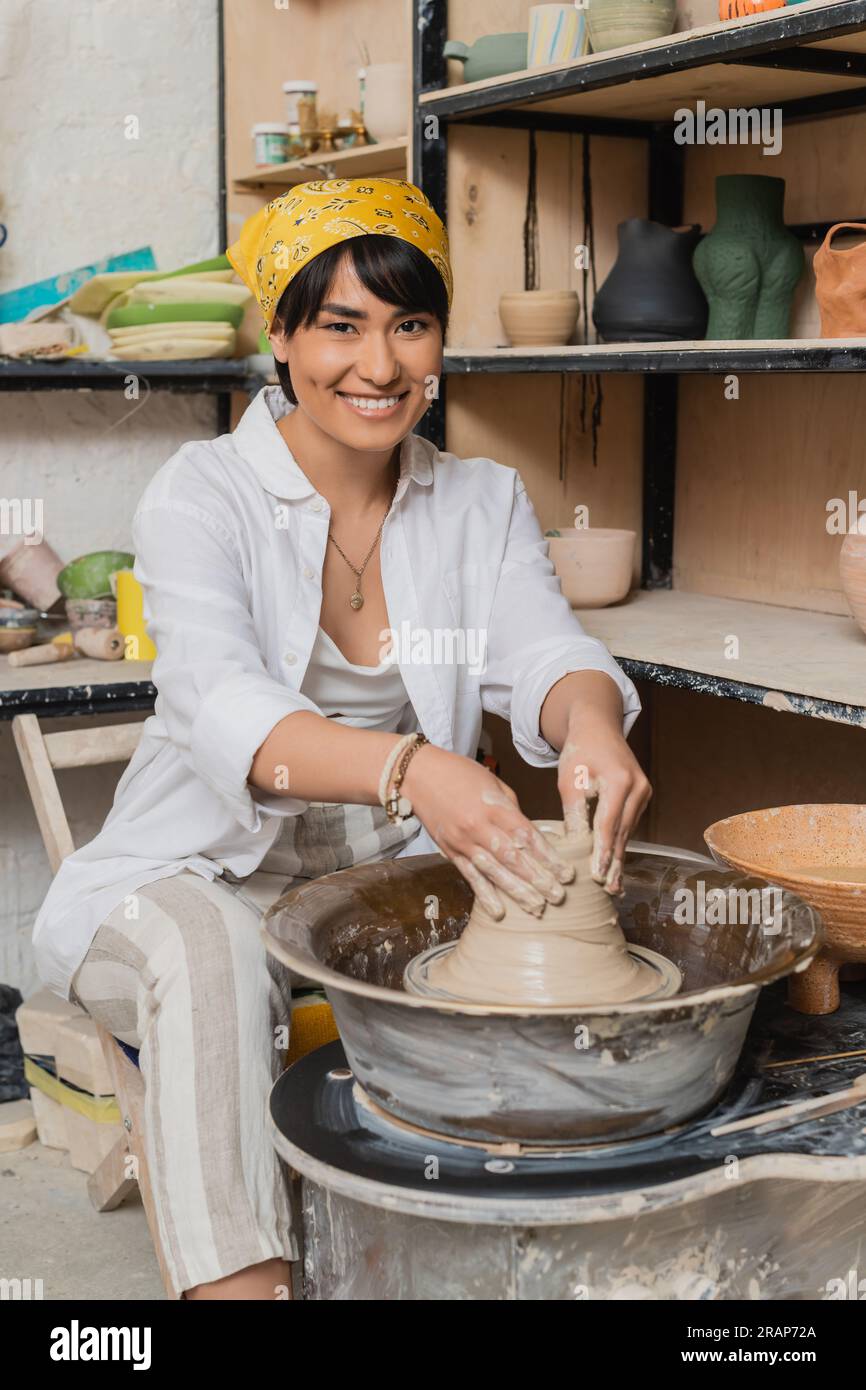Cheerful young asian female artist in headscarf shaping wet clay while ...