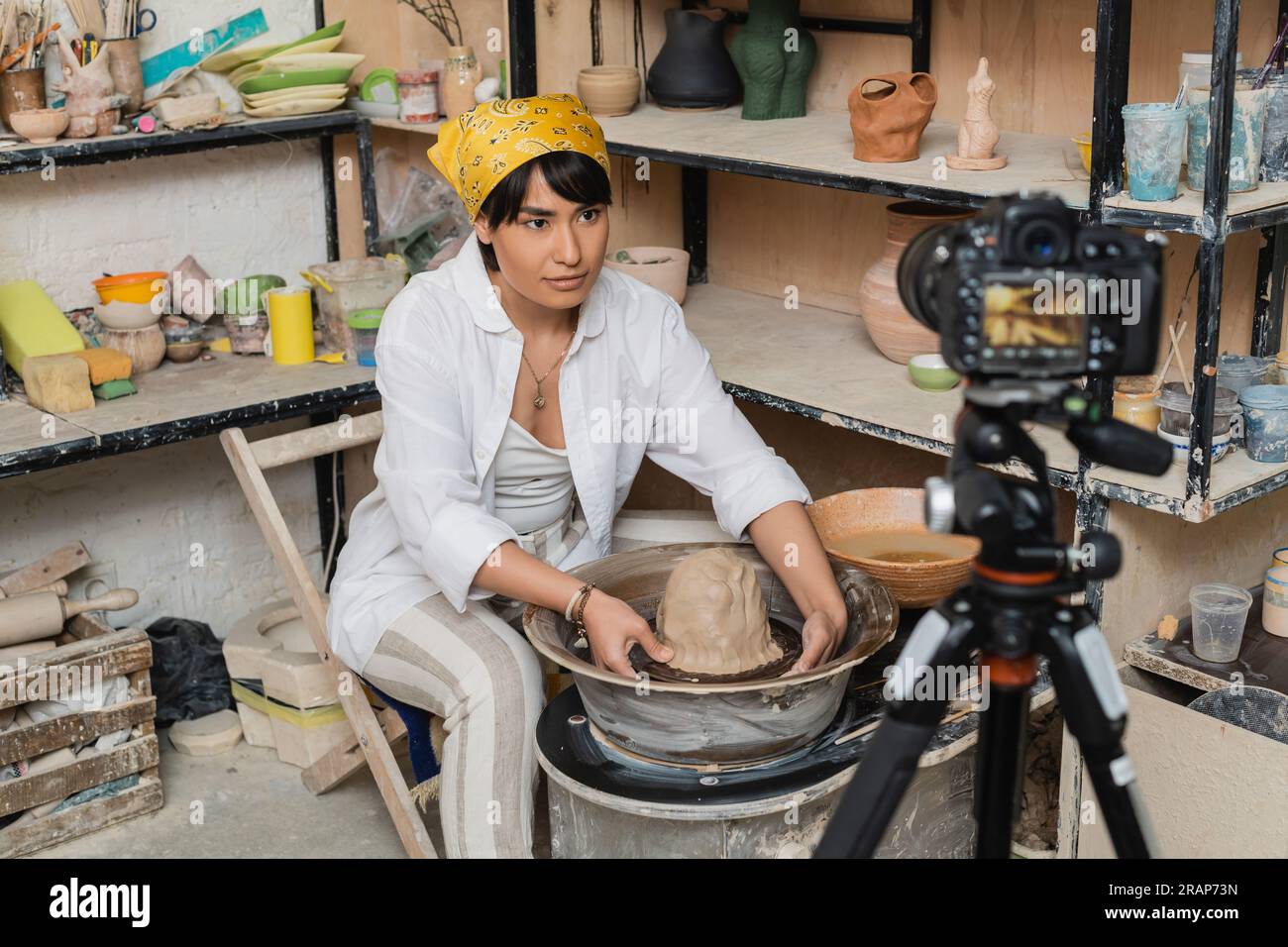 Young asian brunette craftswoman in headscarf and workwear molding clay ...