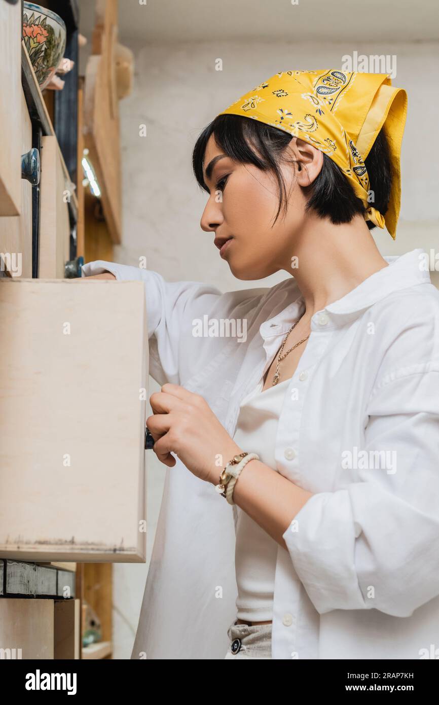 Side view of young brunette asian artist in headscarf and workwear ...