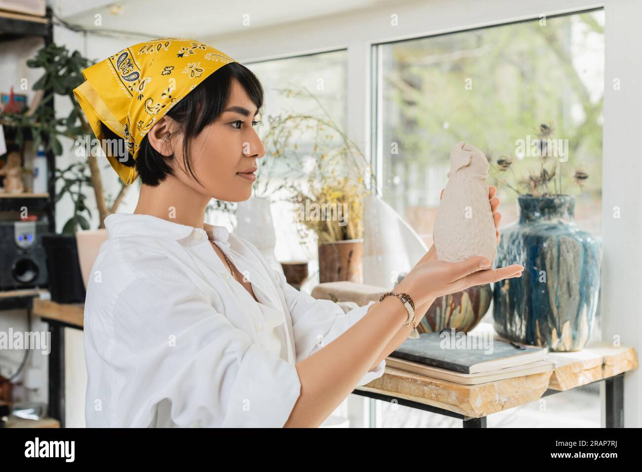 Young female asian artist in workwear and headscarf holding clay ...