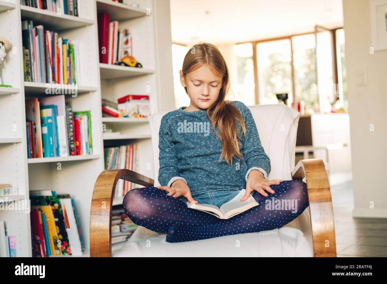Cute little girl sitting in a white chair at home and reading a book ...