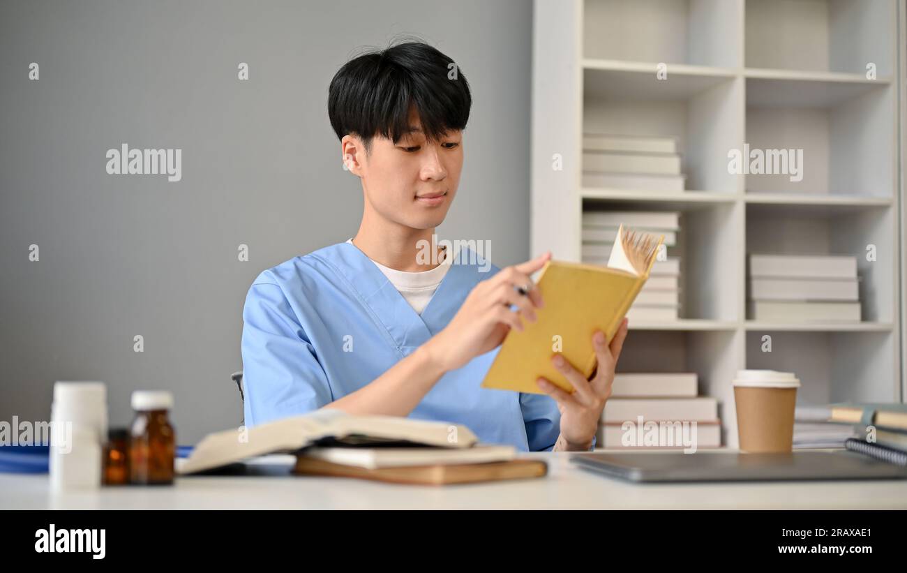 A smart and focused young Asian male medical student in scrubs reading ...