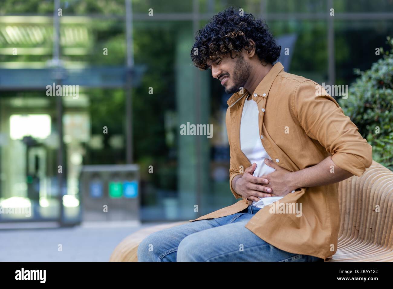 A young Indian man sits outside on a bench and suffers from stomach ...