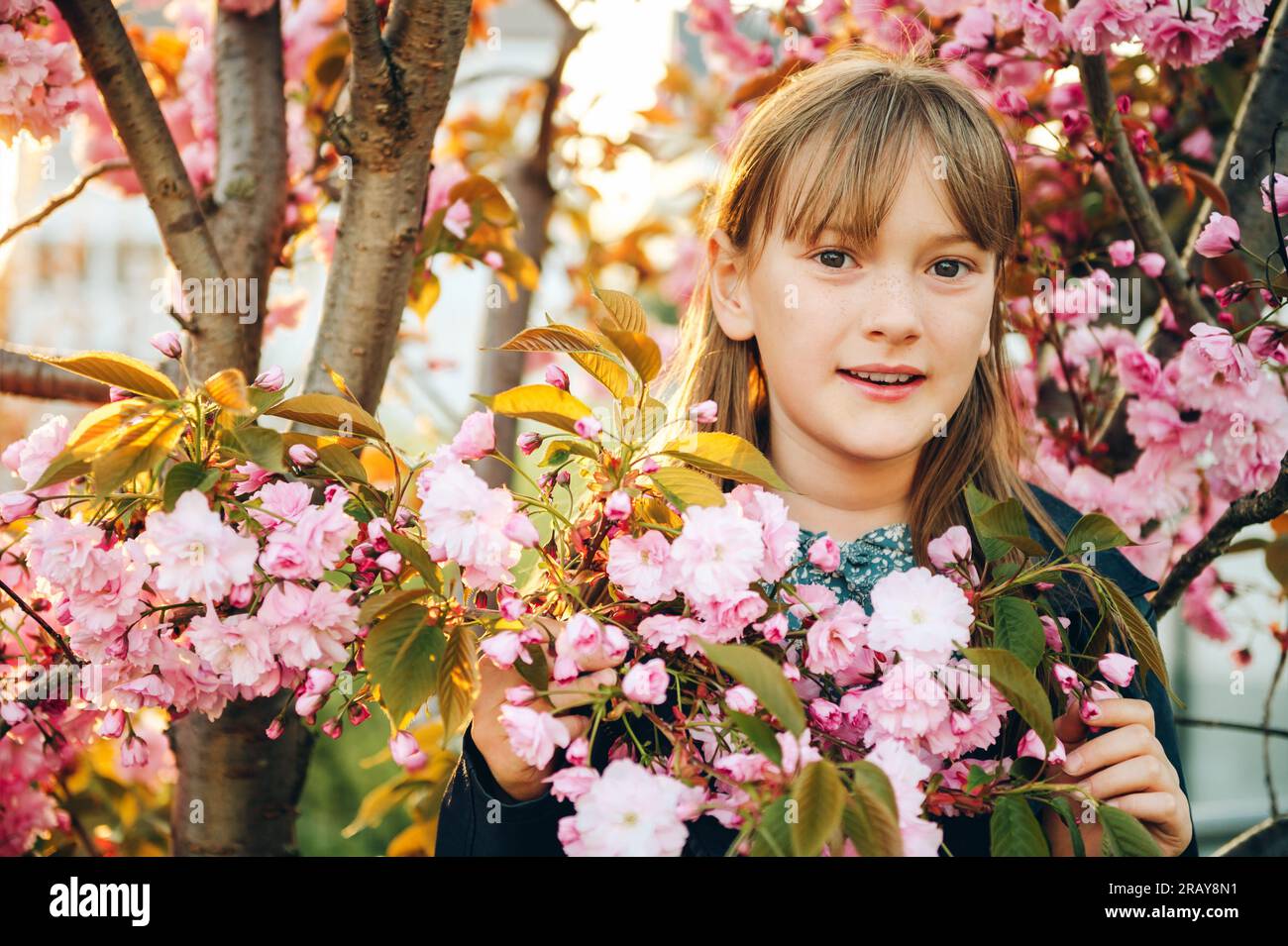 Outdoor spring portrait of a pretty little girl, standing between flowers of a japanese cherry ...