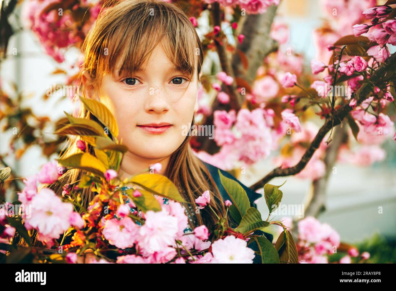 Outdoor spring portrait of a pretty little girl, standing between flowers of a japanese cherry ...