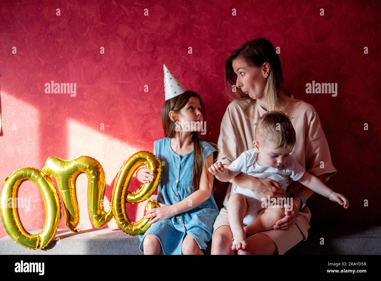 Mother is fooling around on sofa with small children near red wall on gray sofa. Family with one ...