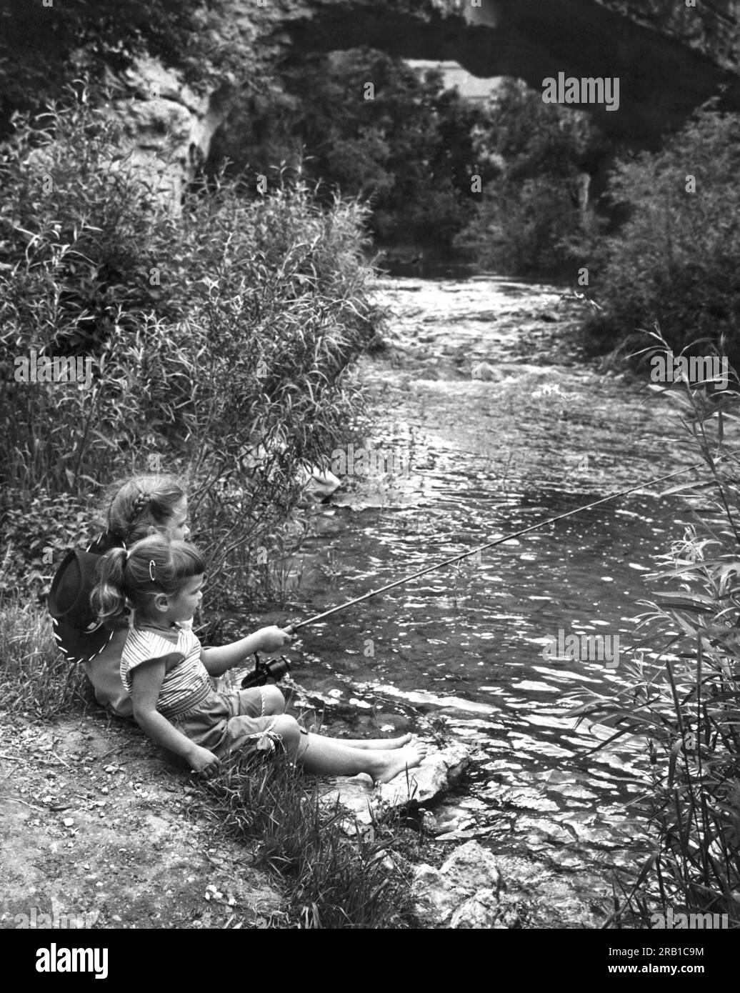 United States: c. 1954 Two young barefoot girls fishing in a small ...