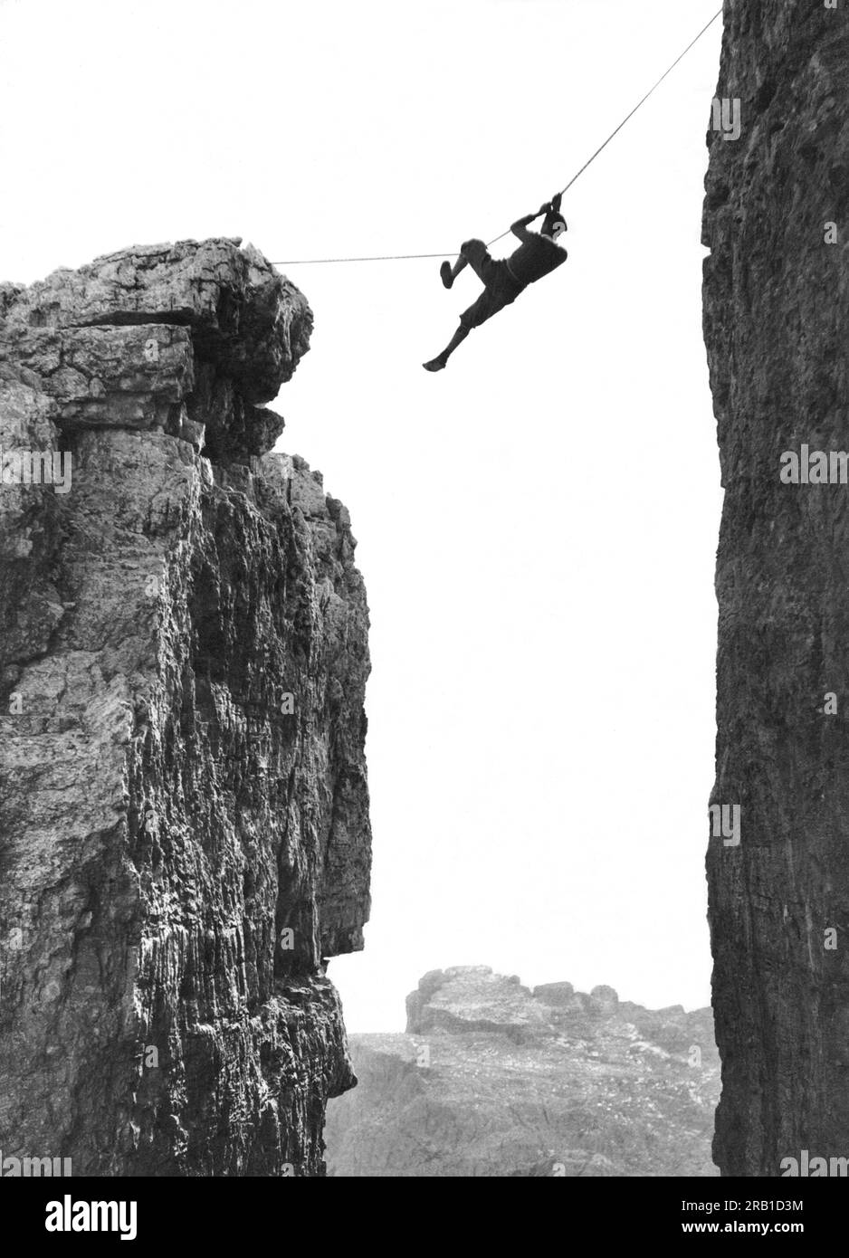 Switzerland: c. 1927 A mountain climber traversing between two cliffs ...