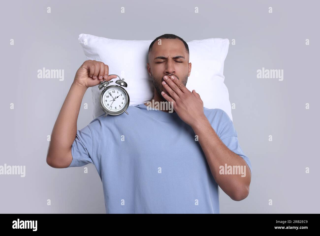 Tired man with pillow and alarm clock yawning on light grey background ...