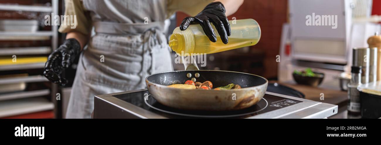The professional chef in uniform adds oil into pan with dish in ...