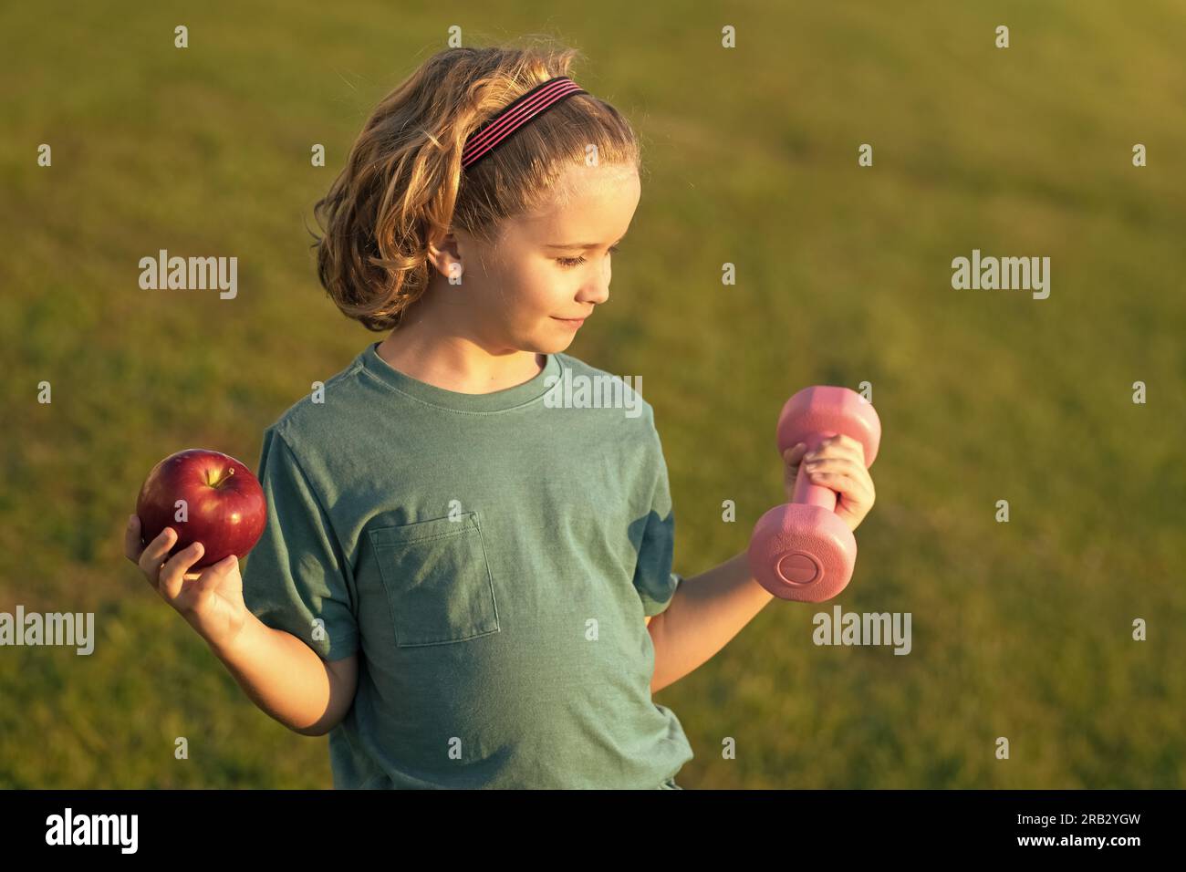 Healthy sporty child with apple and dumbbell outdoor in park. Cute child boy pumping up arm ...