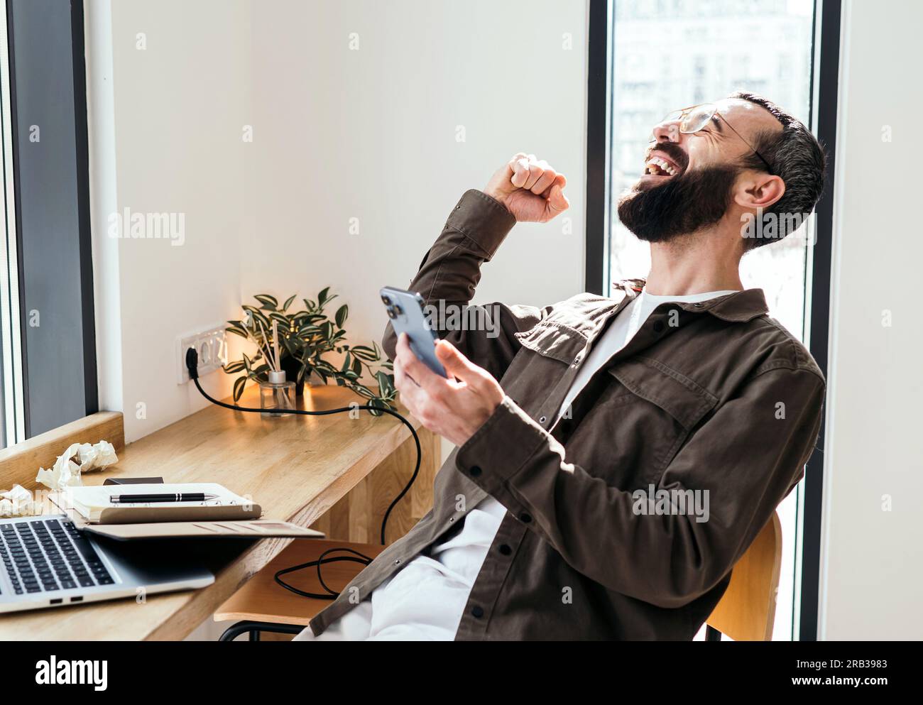 Cheerful bearded man in glasses rejoices in a cafe using a mobile phone ...