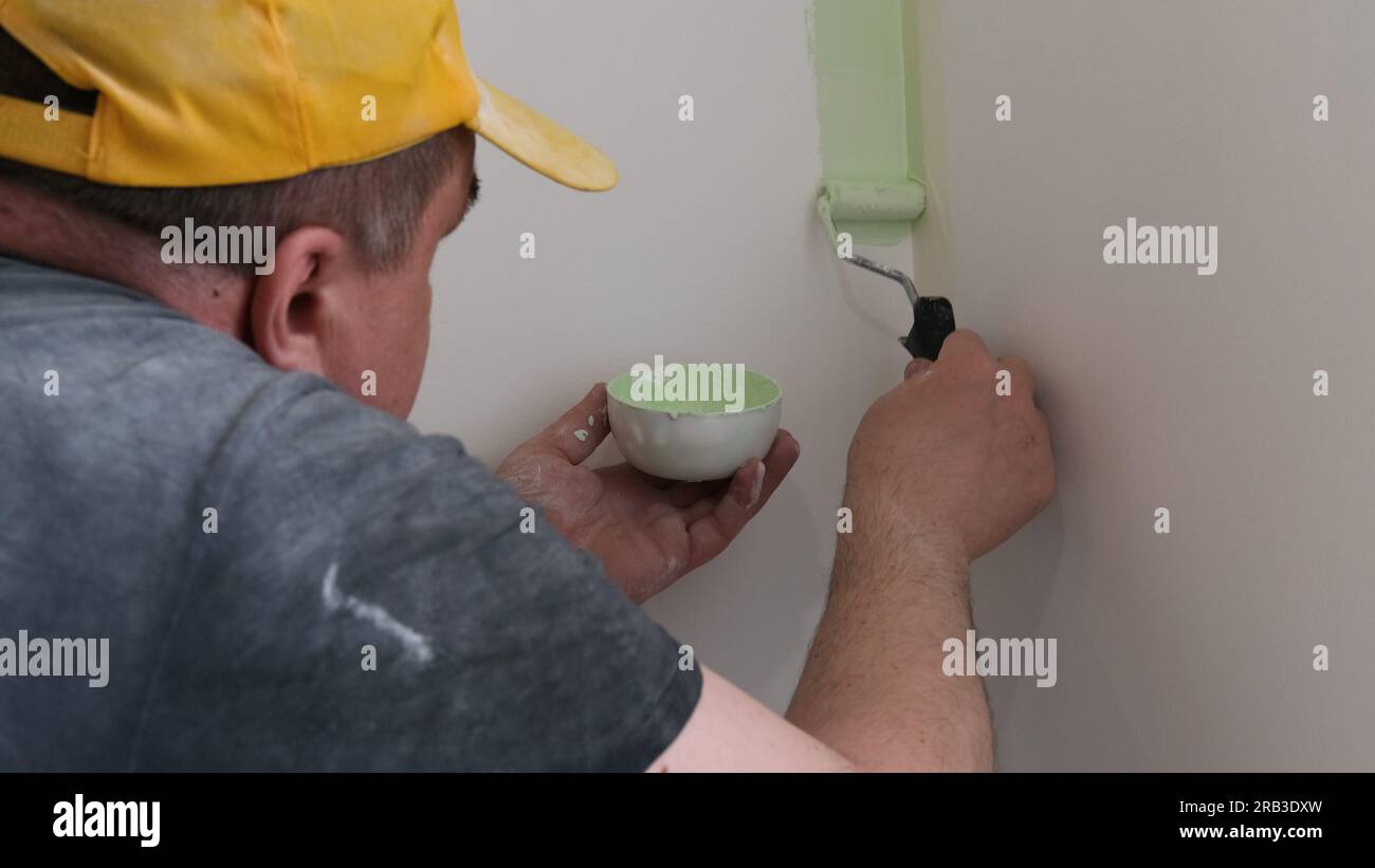 Rear view of a man in a gray T-shirt and cap painting the wall with a ...