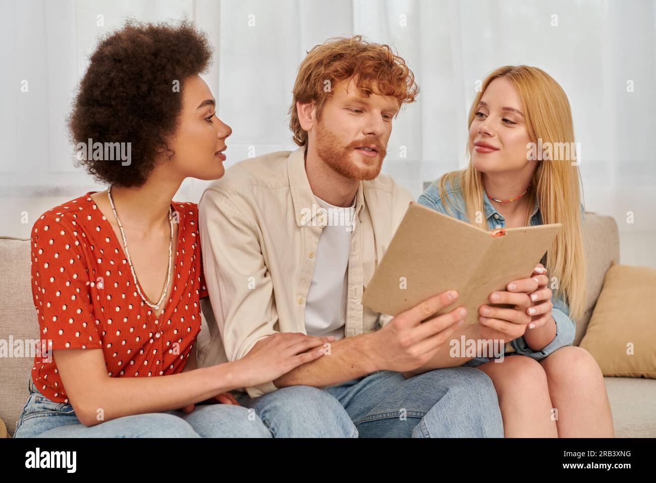 open relationships, redhead man reading book to beautiful multicultural women in living room ...