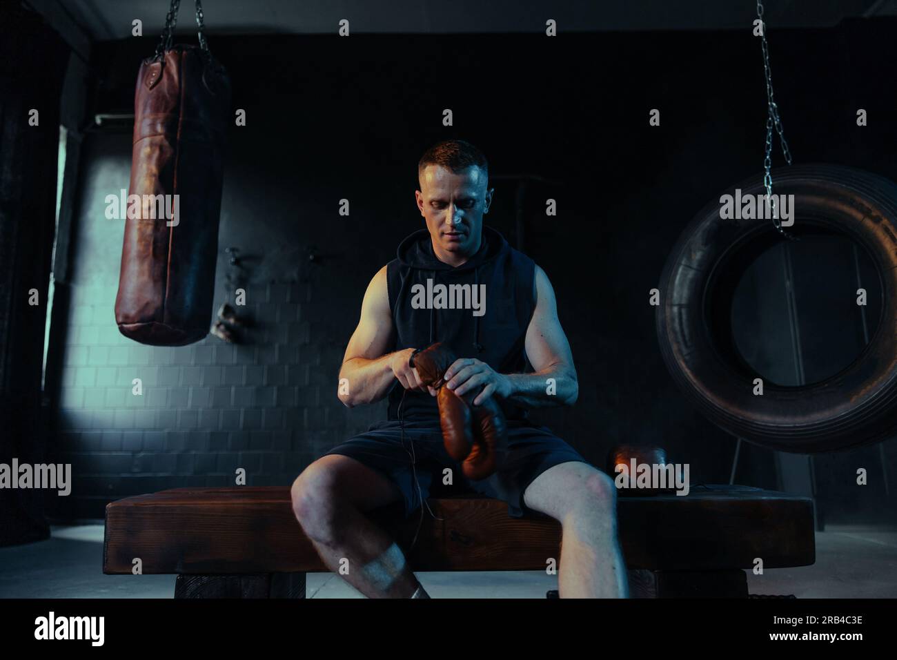 Male boxer sitting on bench in dark gym and putting on boxing gloves ...